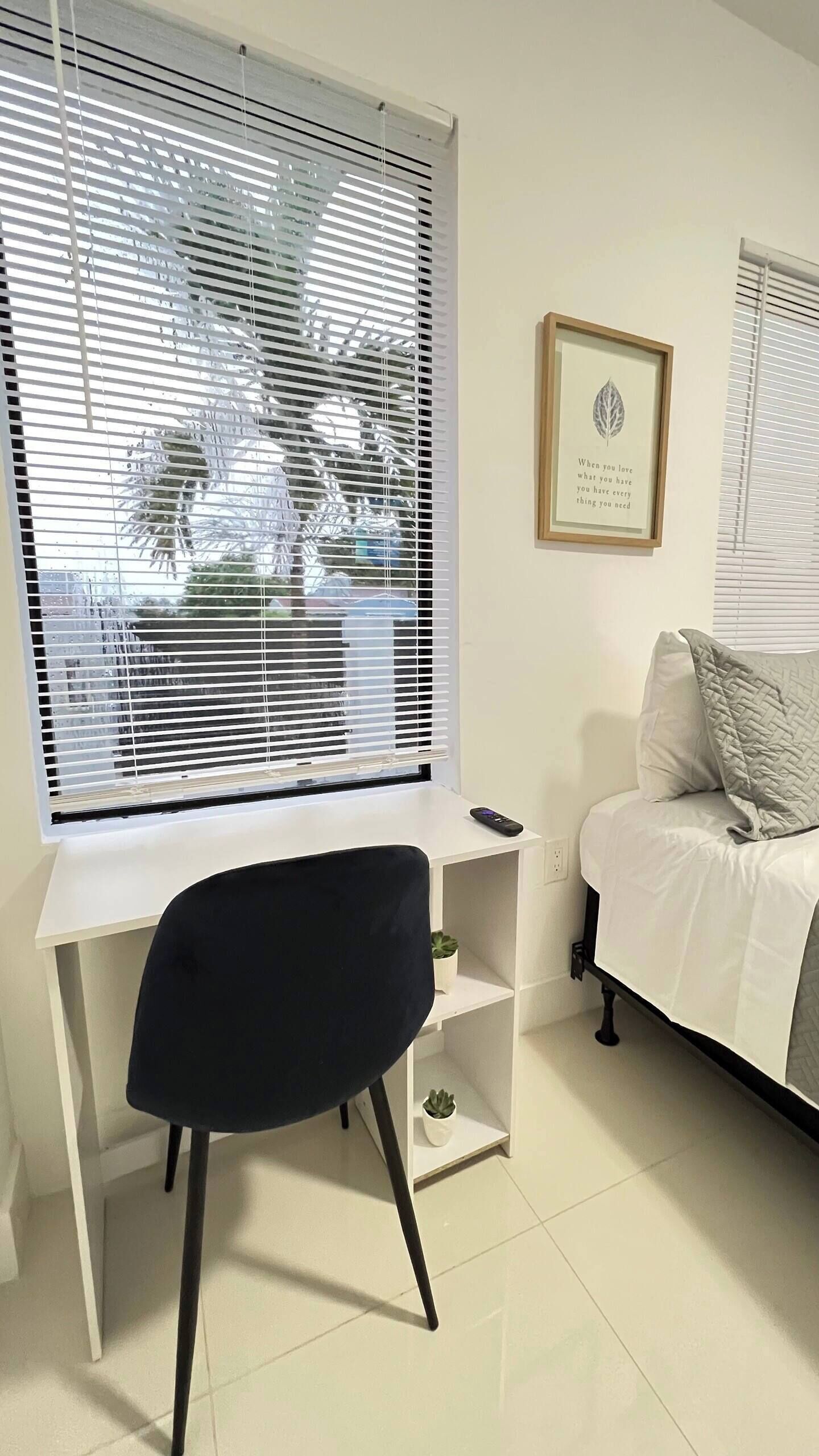 Quarto com cama, escrivaninha com cadeira, uma citação emoldurada na parede e janela com persianas.