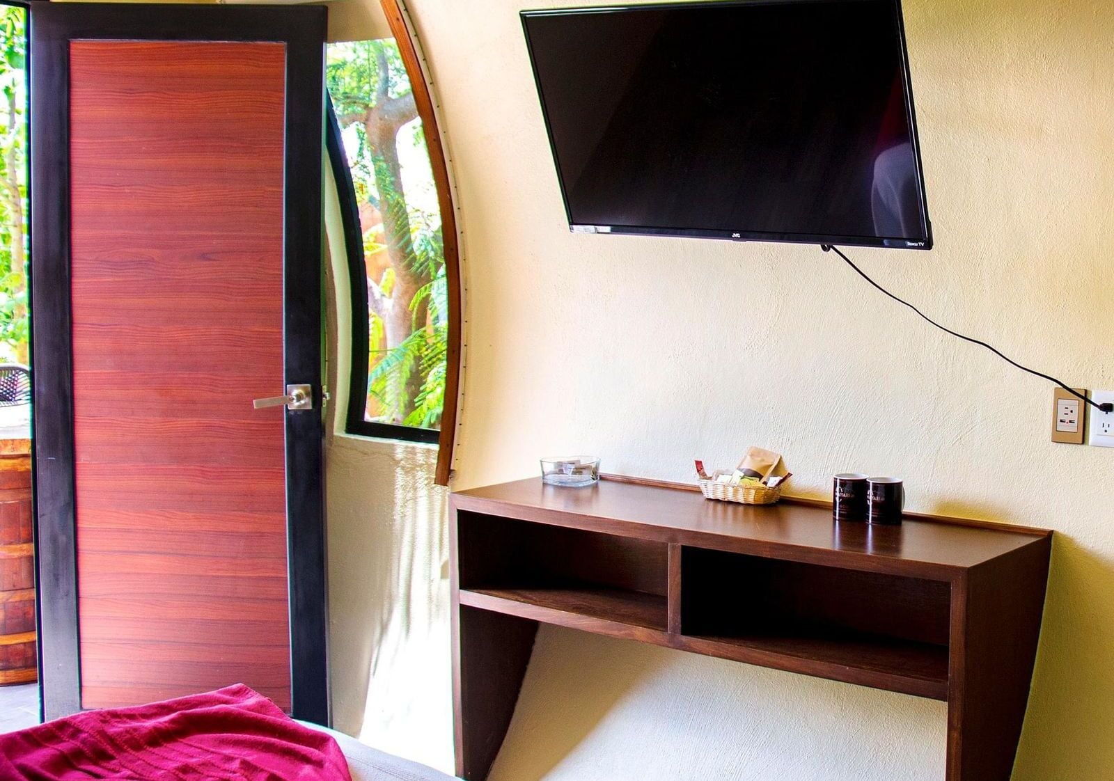 Quarto com televisão na parede, escrivaninha de madeira, cama com manta vermelha e janela com vista para vegetação.