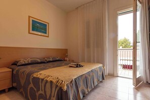 Quarto com cama, mesa de cabeceira, janela com cortinas e vista para varanda.