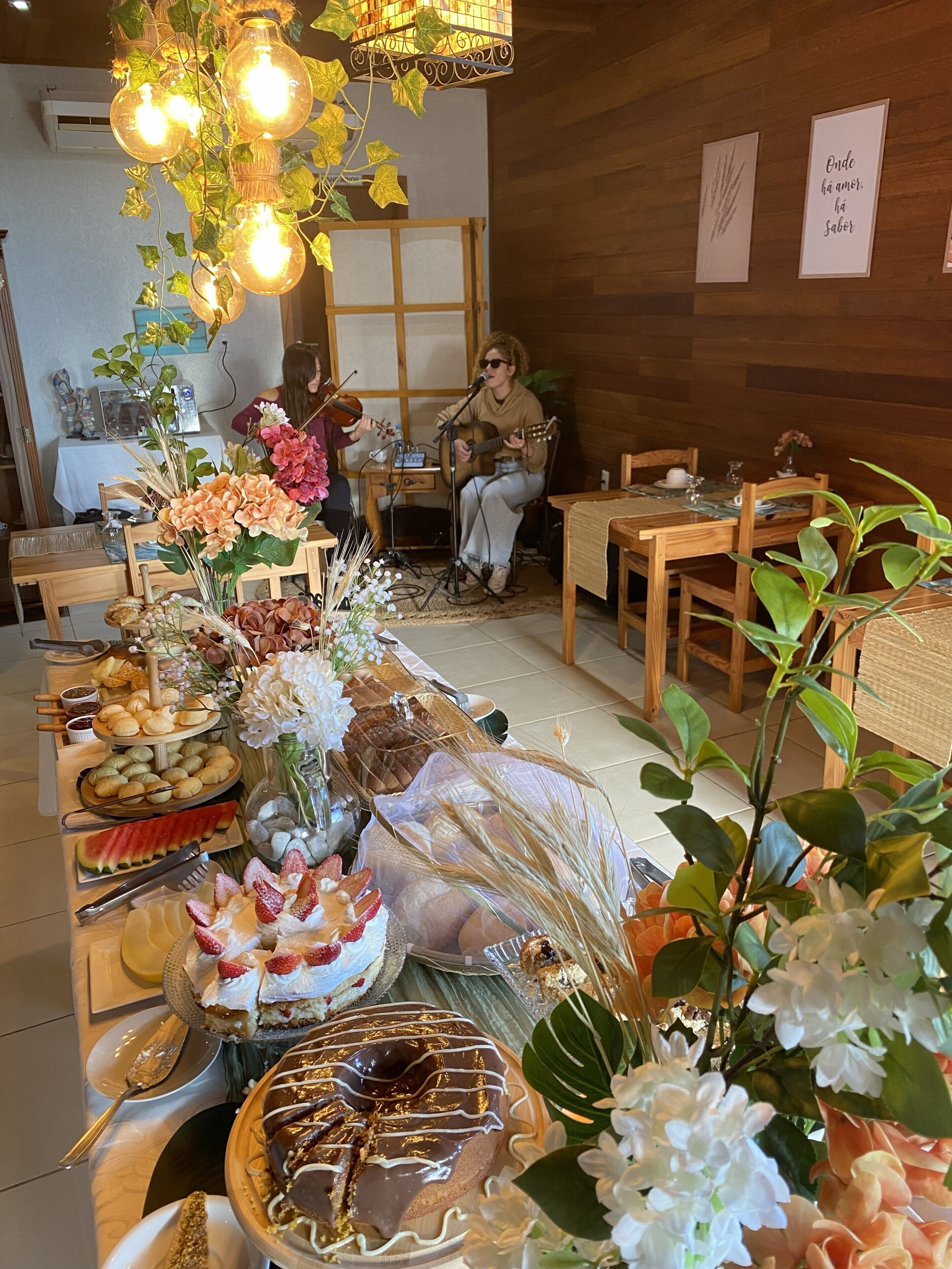 Uma sala de jantar com uma mesa comprida posta para refeição, um músico tocando violão e um lustre decorativo.
