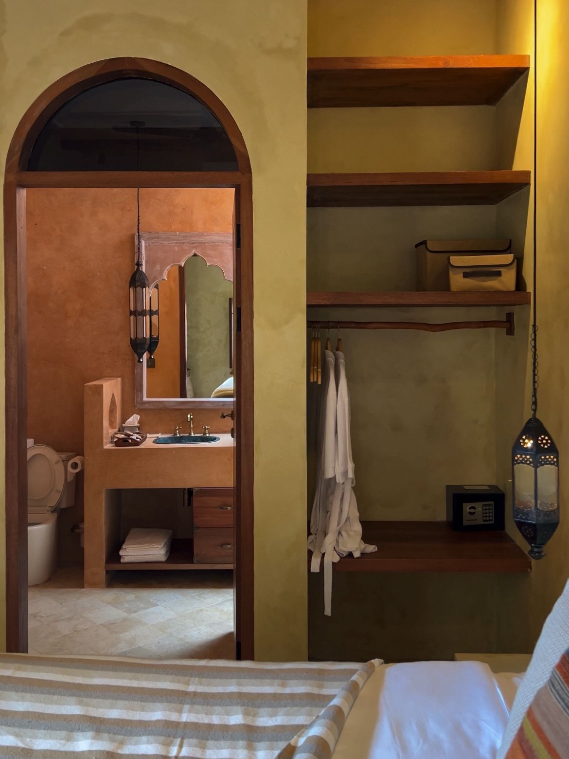 Quarto com uma cama, porta em arco de madeira dando para um banheiro, prateleira com uma caixa e um candeeiro pendente.