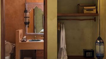 Quarto com uma cama, porta em arco de madeira dando para um banheiro, prateleira com uma caixa e um candeeiro pendente.