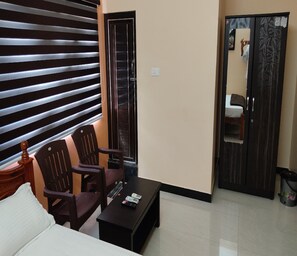 Quarto com sofá, duas cadeiras de madeira, mesa de centro com controles remotos, porta com espelho e lixeira.