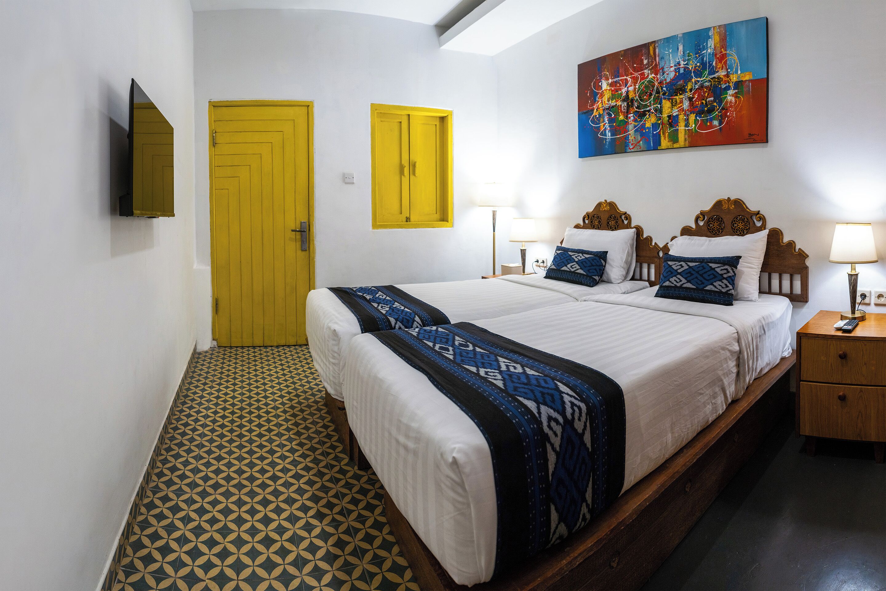 Quarto de hotel com uma cama grande, duas mesas de cabeceira, uma TV fixada na parede e um quadro abstrato colorido sobre a cama.