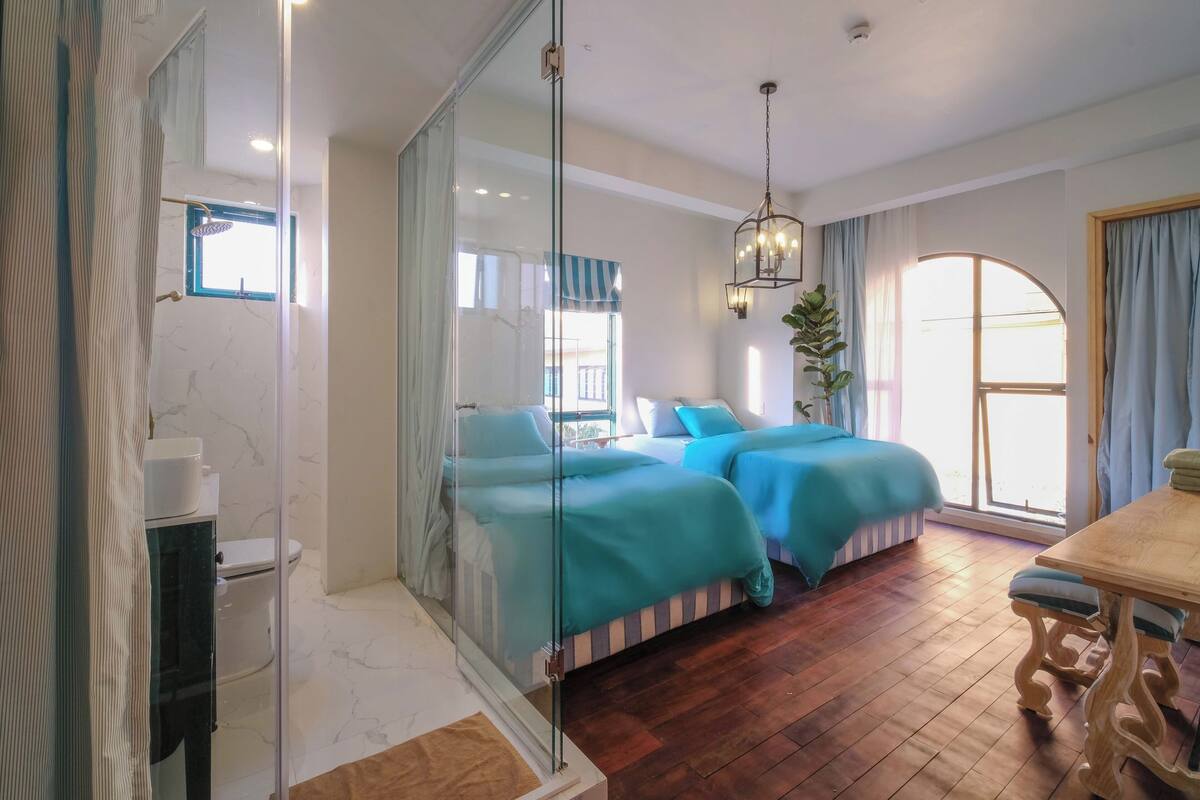 Quarto com duas camas, piso de madeira, divisória de vidro, banheiro com chuveiro e uma janela grande.