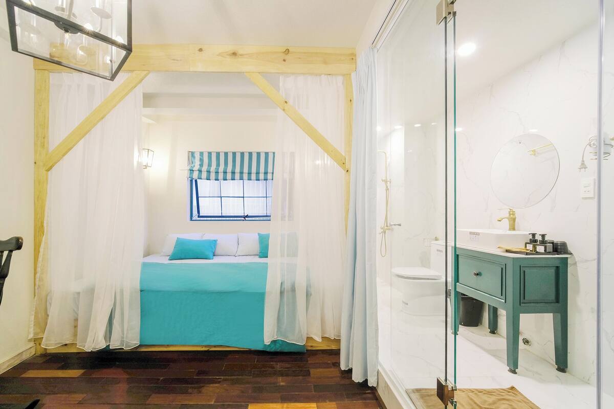 Quarto com cama, janela, banheiro com pia e espelho, e estrutura de teto de madeira.