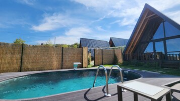 Piscina com deck de madeira e espreguiçadeiras, cercada por um cercado de bambu e um edifício moderno com telhado inclinado.