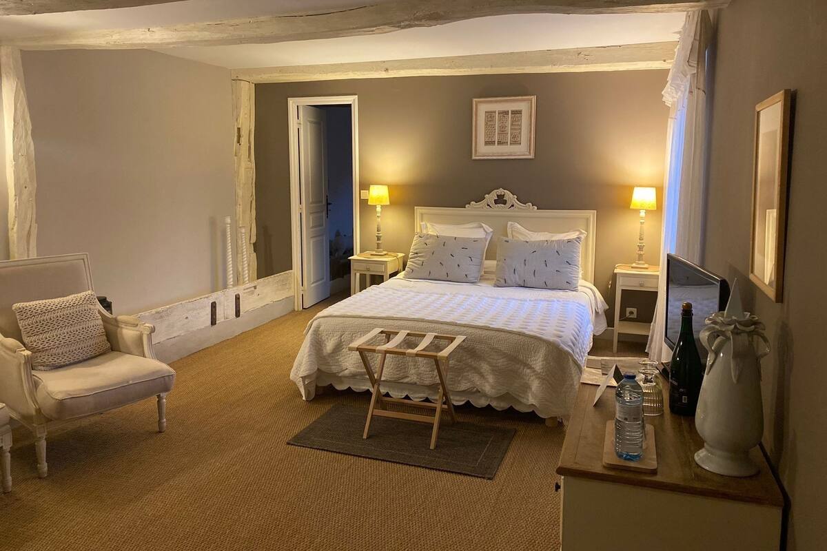 Quarto com cama, mesas de cabeceira, uma cadeira, um banquinho e uma escrivaninha com uma garrafa e copos.