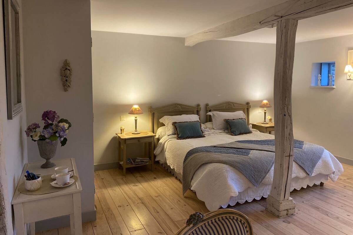 Quarto com cama grande, duas mesas de cabeceira com abajures, uma mesinha com um vaso de flores e uma cadeira de madeira.