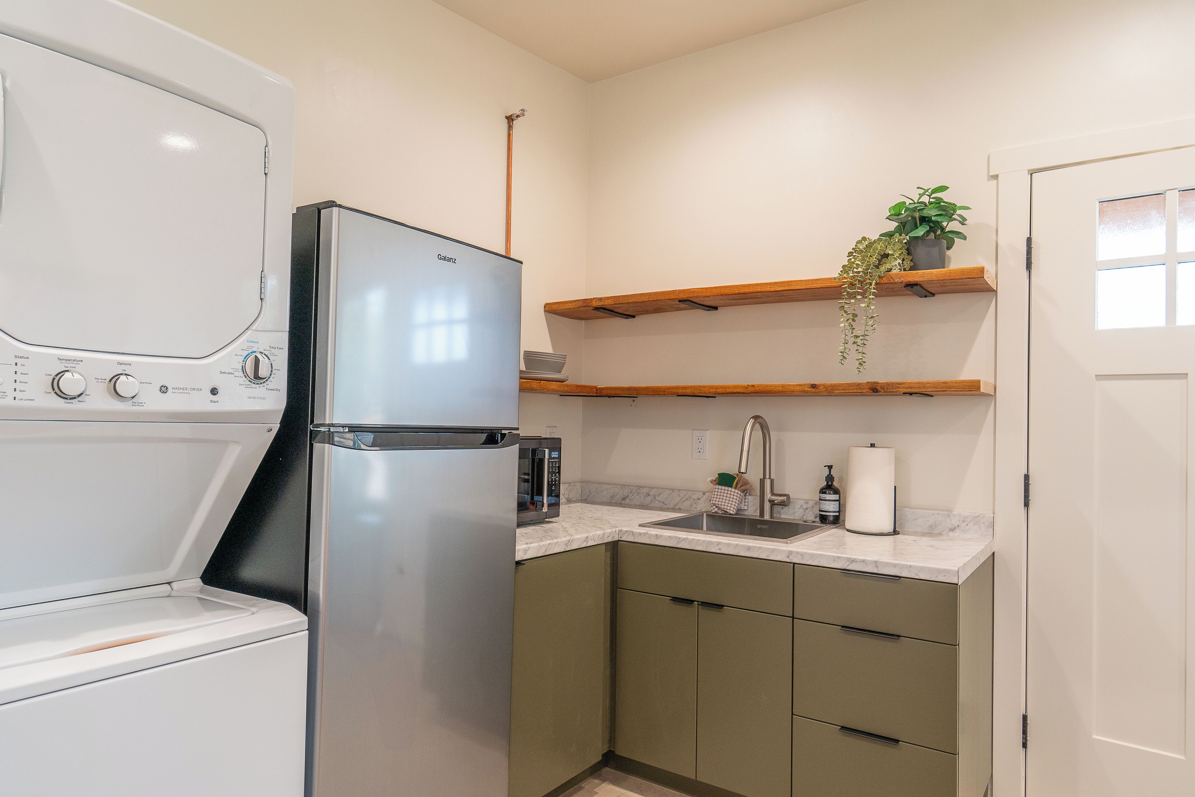 Cozinha compacta com máquina de lavar e secar embutidas, frigorífico, micro-ondas e lava-loiça com estantes de madeira acima.