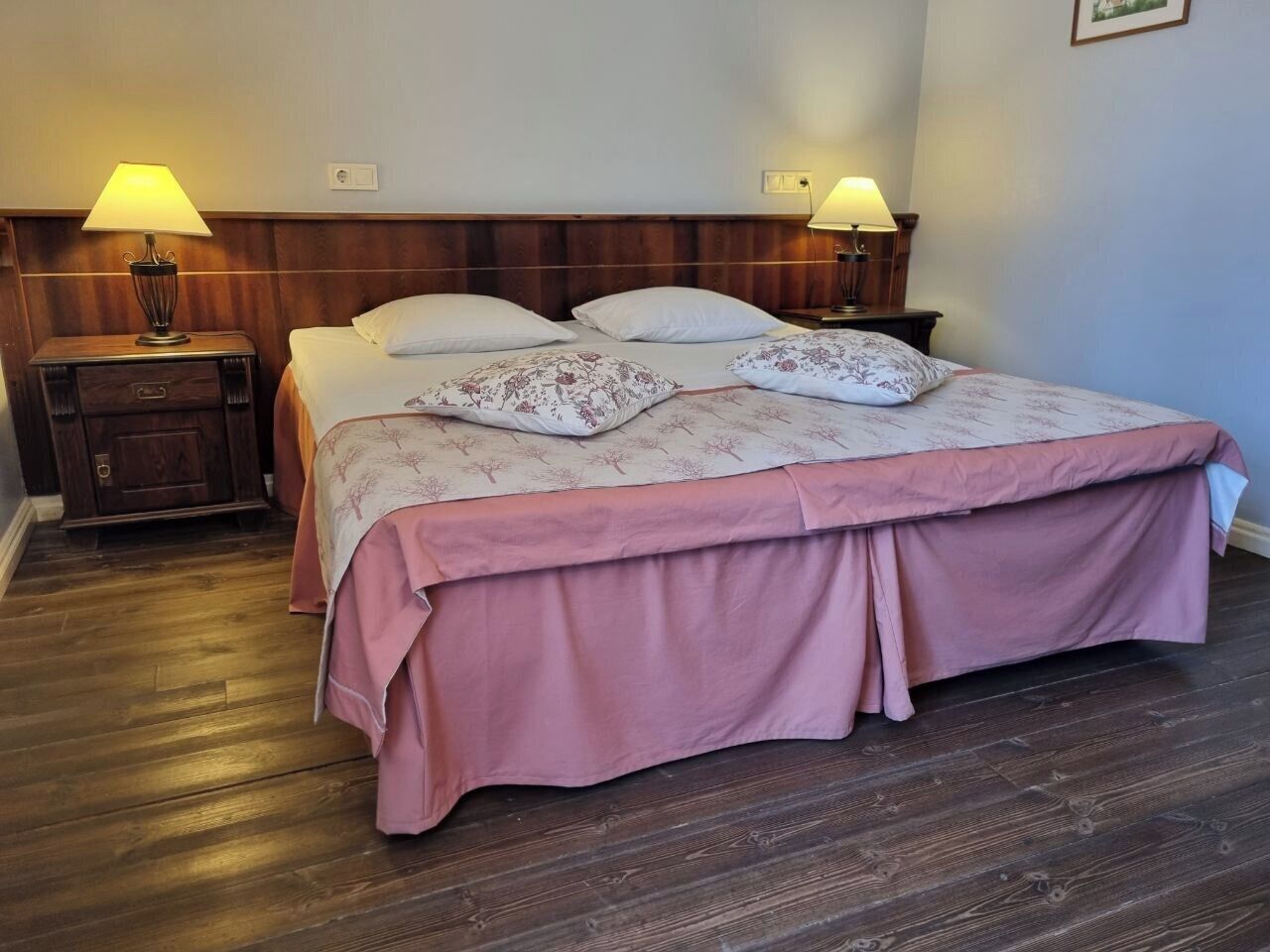 Uma cama de casal com saia rosa, duas luminárias de cabeceira e um painel de cabeceira de madeira.