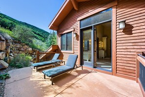 Uma cabana de madeira com varanda, duas poltronas, uma parede de pedra e vista para uma encosta arborizada.