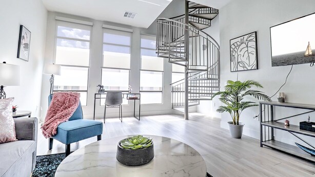 Uma sala de estar moderna com uma poltrona azul, uma mesa de centro redonda com uma planta, uma escada em espiral e janelas amplas que oferecem vista do céu.