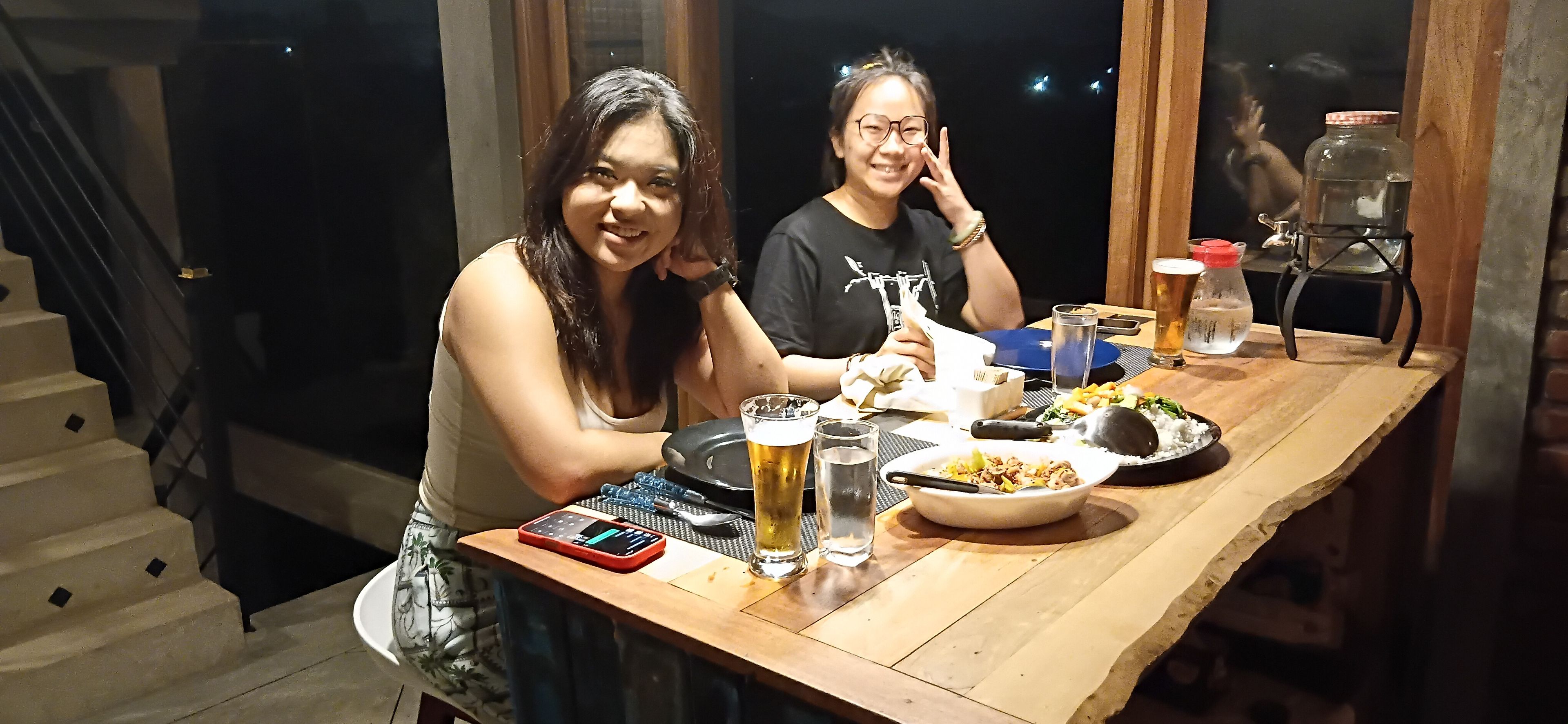 Duas pessoas sentadas numa mesa de madeira, com comida, bebidas e um smartphone.
