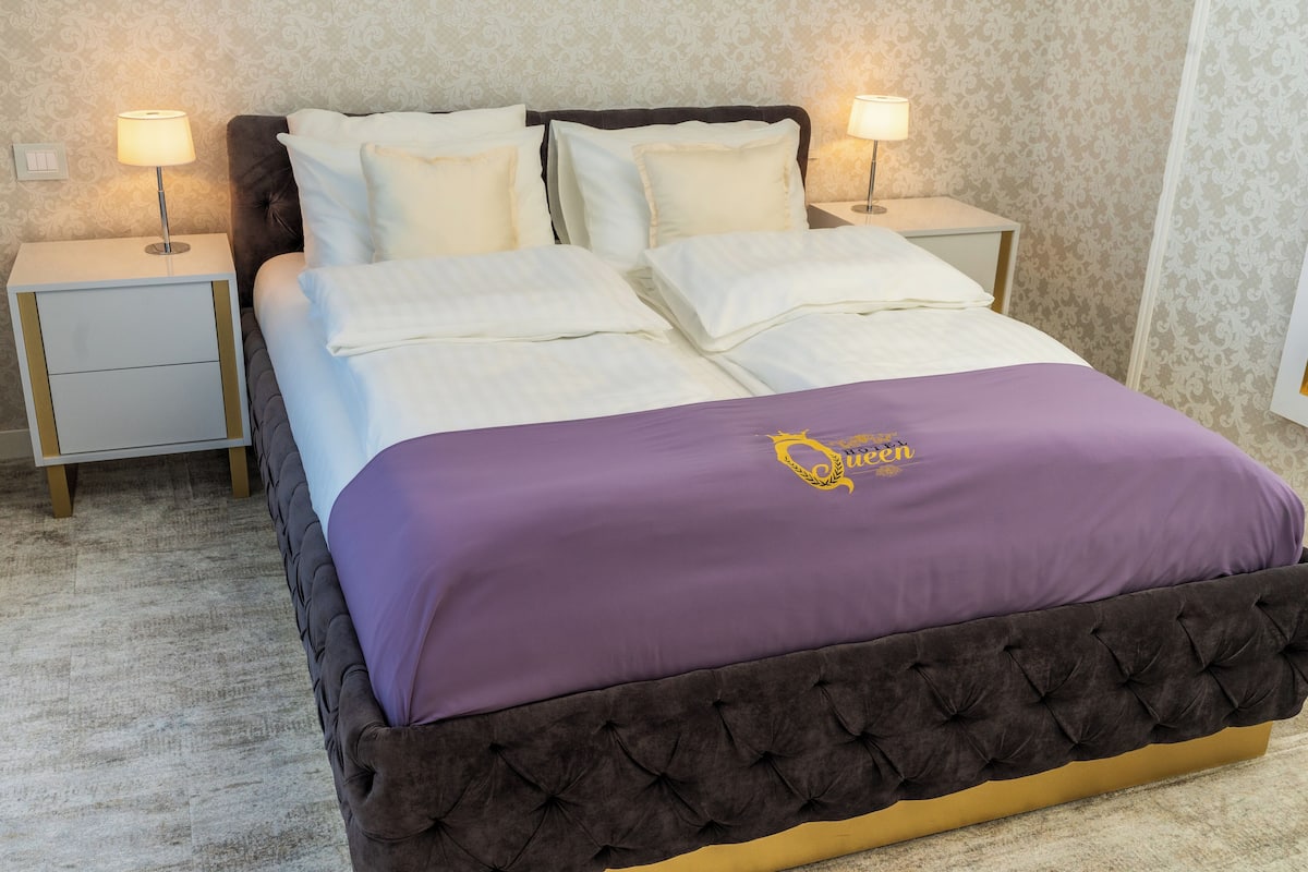 Cama bem arrumada com colcha roxa e um logotipo, ladeada por duas mesas de cabeceira com abajures, em quarto com papel de parede estampado.