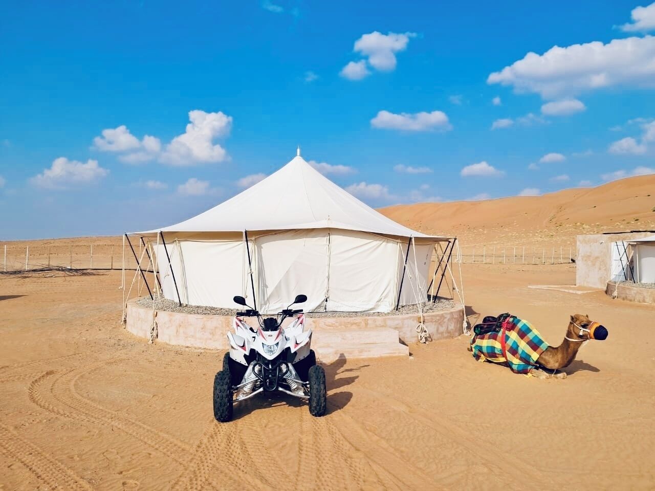 Cena de deserto com uma tenda branca, um quadriciclo e um camelo.