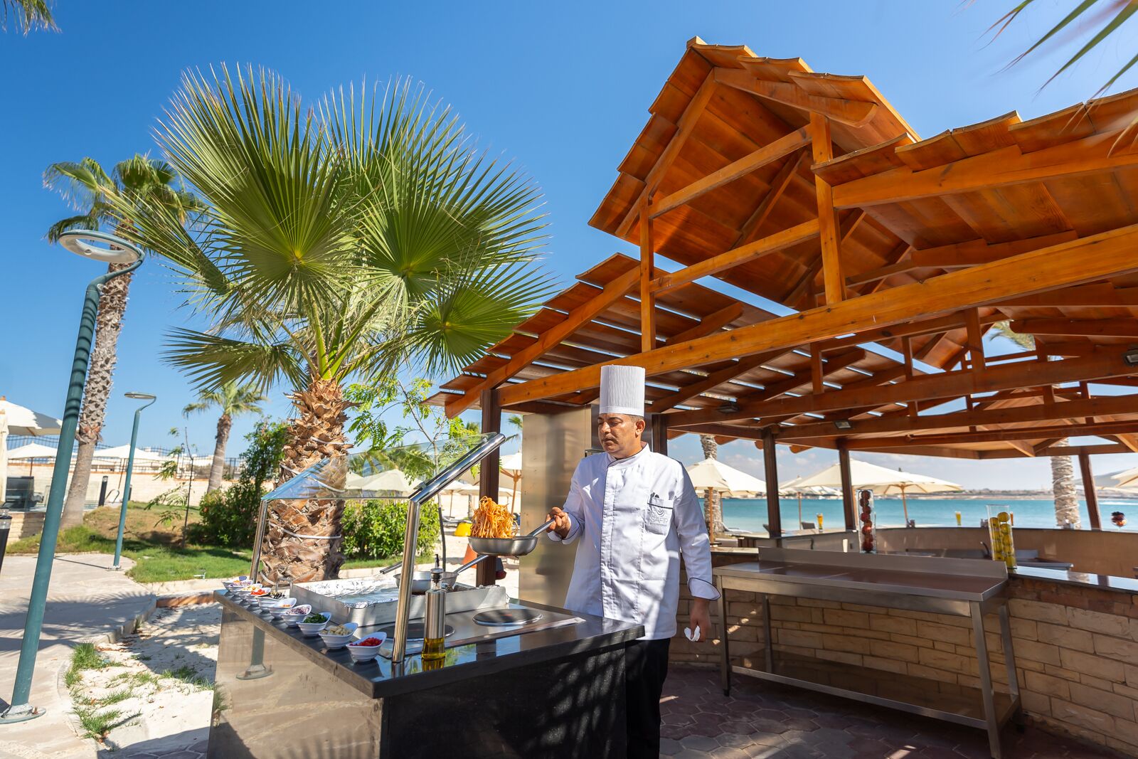 Um chef preparando comida ao ar livre, com uma pérgola de madeira e palmeiras ao fundo.