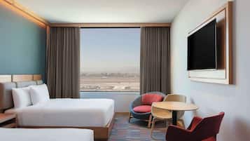 Quarto de hotel com uma cama grande, uma escrivaninha, duas cadeiras, uma televisão e uma janela com vista para um aeroporto.