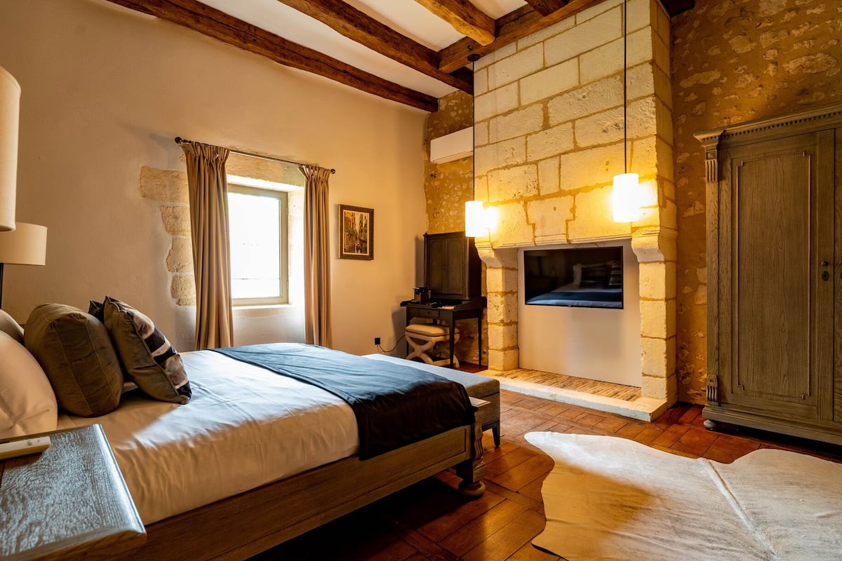Quarto com cama, lareira, parede de pedra e vigas de madeira.