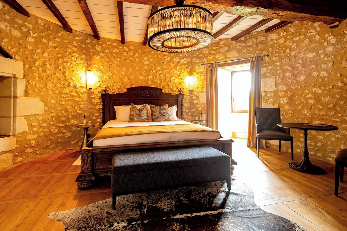 Quarto com uma cama grande, um candelabro, uma parede de pedra, piso de madeira e tapete de couro.