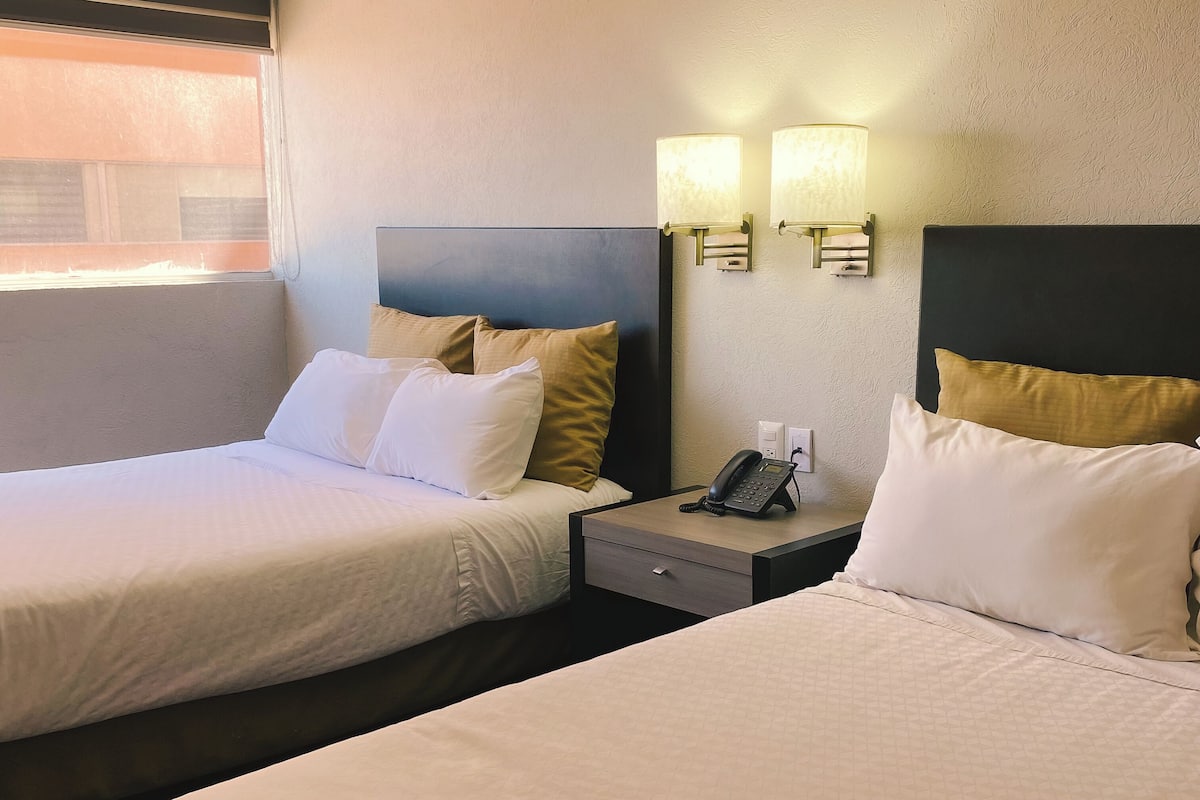 Quarto de hotel com duas camas, mesa de cabeceira com telefone e luminárias fixadas na parede.