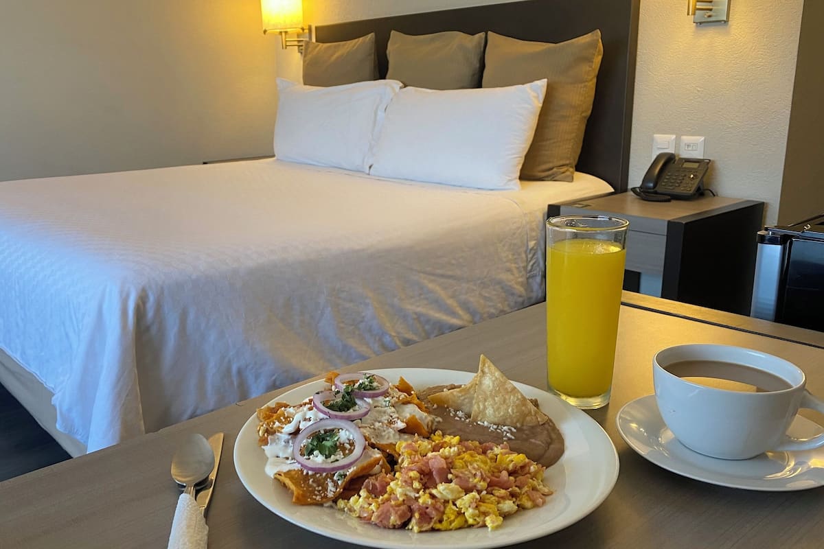 Quarto de hotel com cama bem arrumada, uma mesa de café da manhã com um prato de comida, um copo de suco de laranja e uma xícara de café.