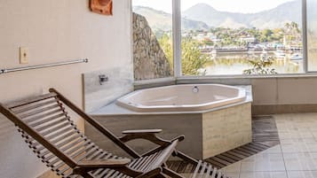 Um banheiro com banheira, cadeiras de madeira, parede de pedra e vista para um lago e montanhas.