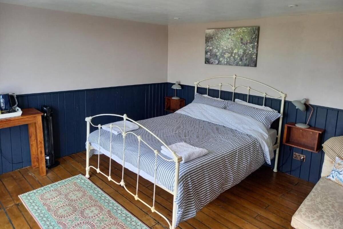 Quarto com estrutura de cama de metal, mesa de cabeceira de madeira, pequena luminária de mesa e uma obra de arte na parede.