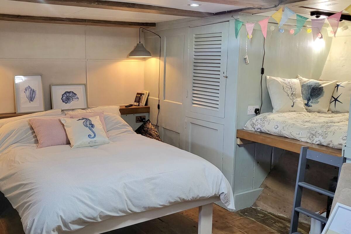 Um quarto com uma cama grande, uma cama menor, uma escrivaninha e uma escada. Há quadros nas paredes e uma prateleira com livros e um abajur.