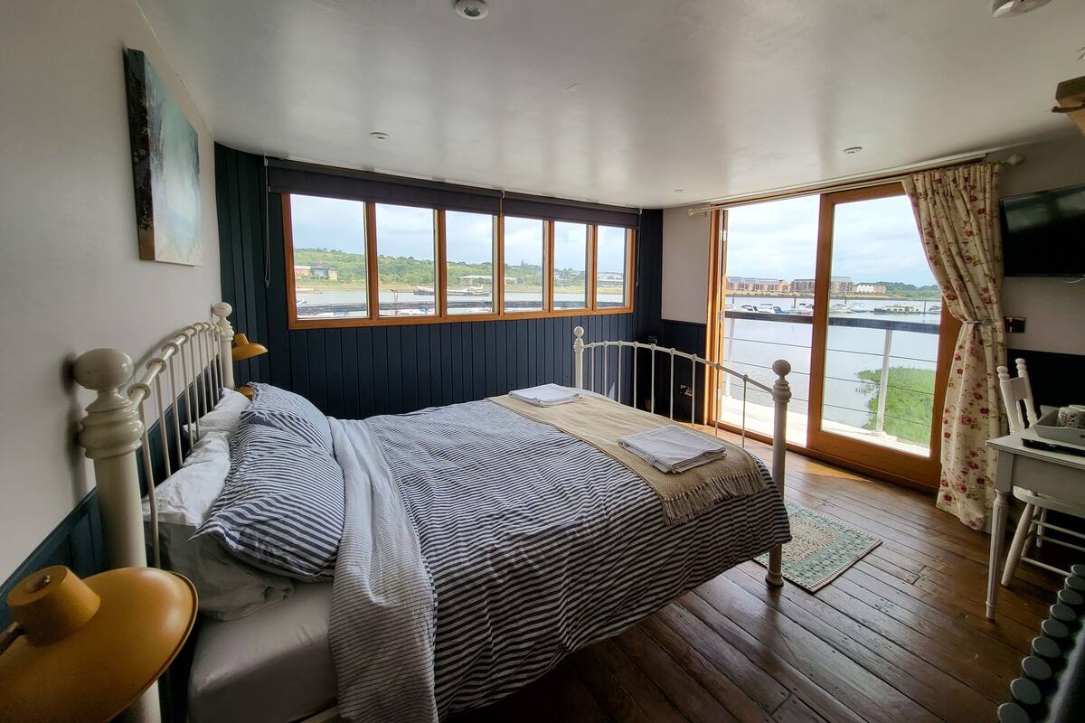 Quarto com cama, escrivaninha e vista para um corpo d'água através de amplas janelas.