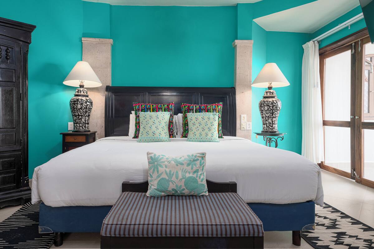 Quarto com uma cama grande, duas mesas de cabeceira, um banquinho e uma janela com cortinas.