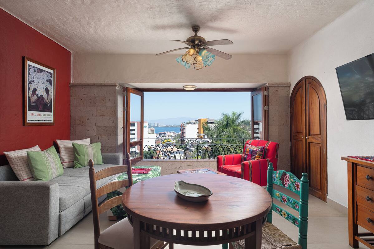 Quarto com varanda, ventoinha no teto, sofá, mesa de jantar e armário de madeira.