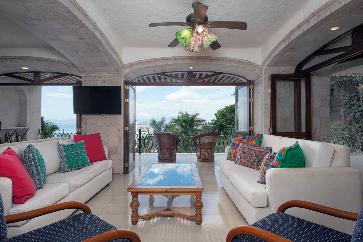 Ampla sala de estar com ventoinha no teto, dois sofás com almofadas coloridas e uma mesa de centro de madeira, além de vista para o exterior.