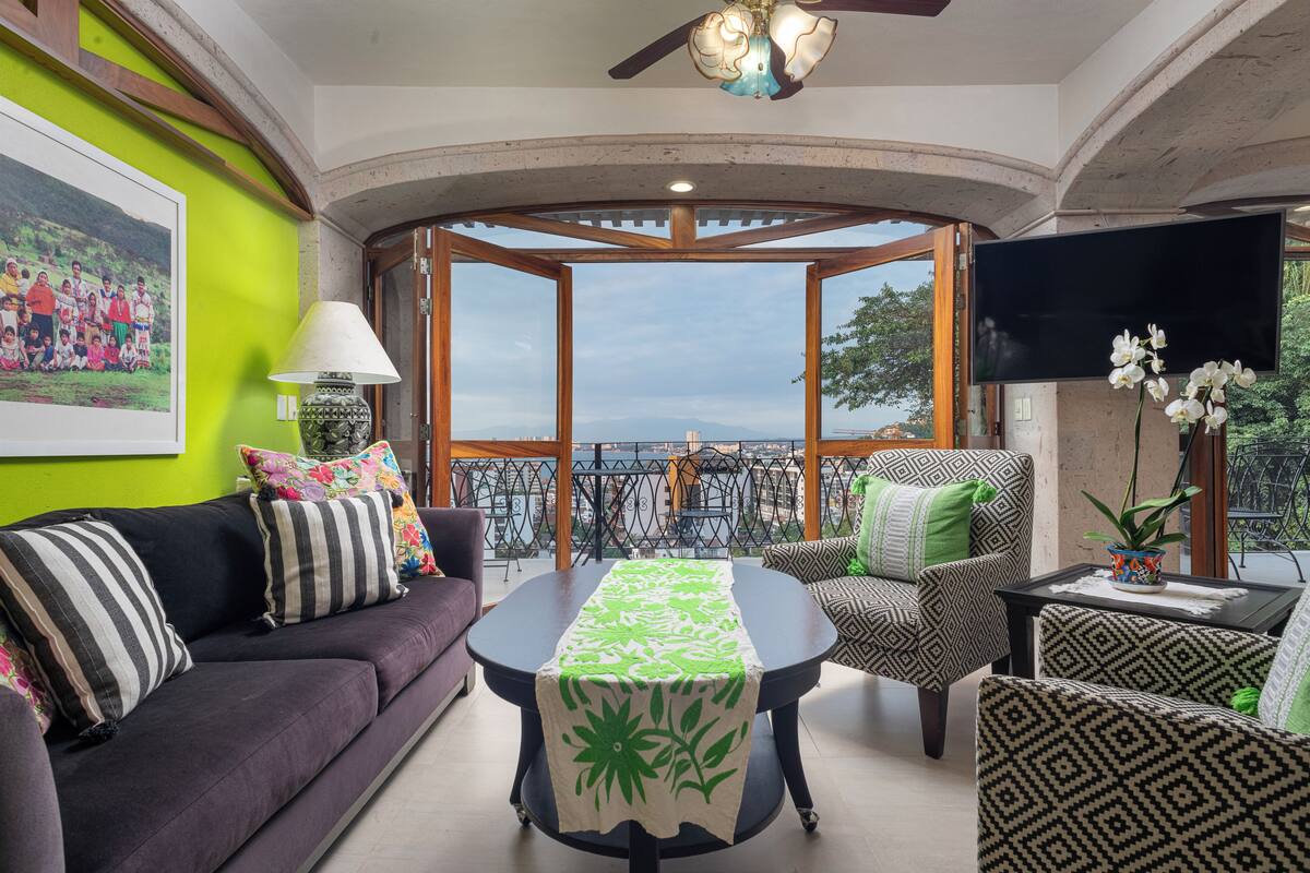 Quarto com sofá, cadeiras, mesa com toalha verde, televisão e varanda com vista.