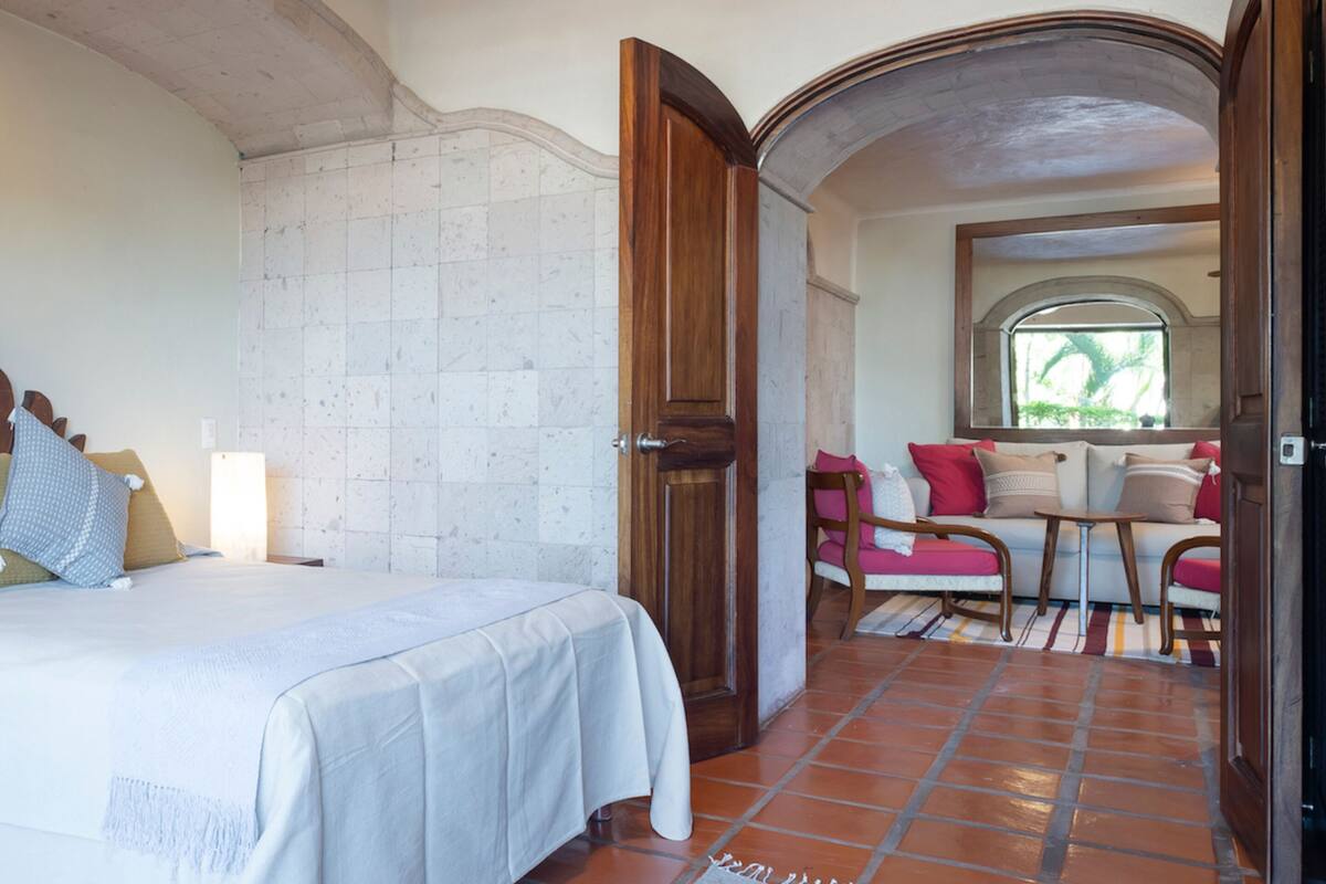 Quarto com cama grande, cabeceira de madeira, mesa de cabeceira e porta que dá para um espaço de estar com sofá e cadeiras.