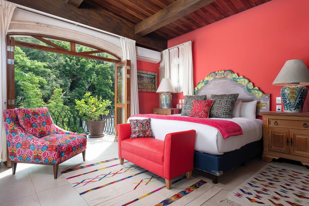 Quarto com uma cama grande, uma cadeira, uma mesa de cabeceira e vista para o verde.