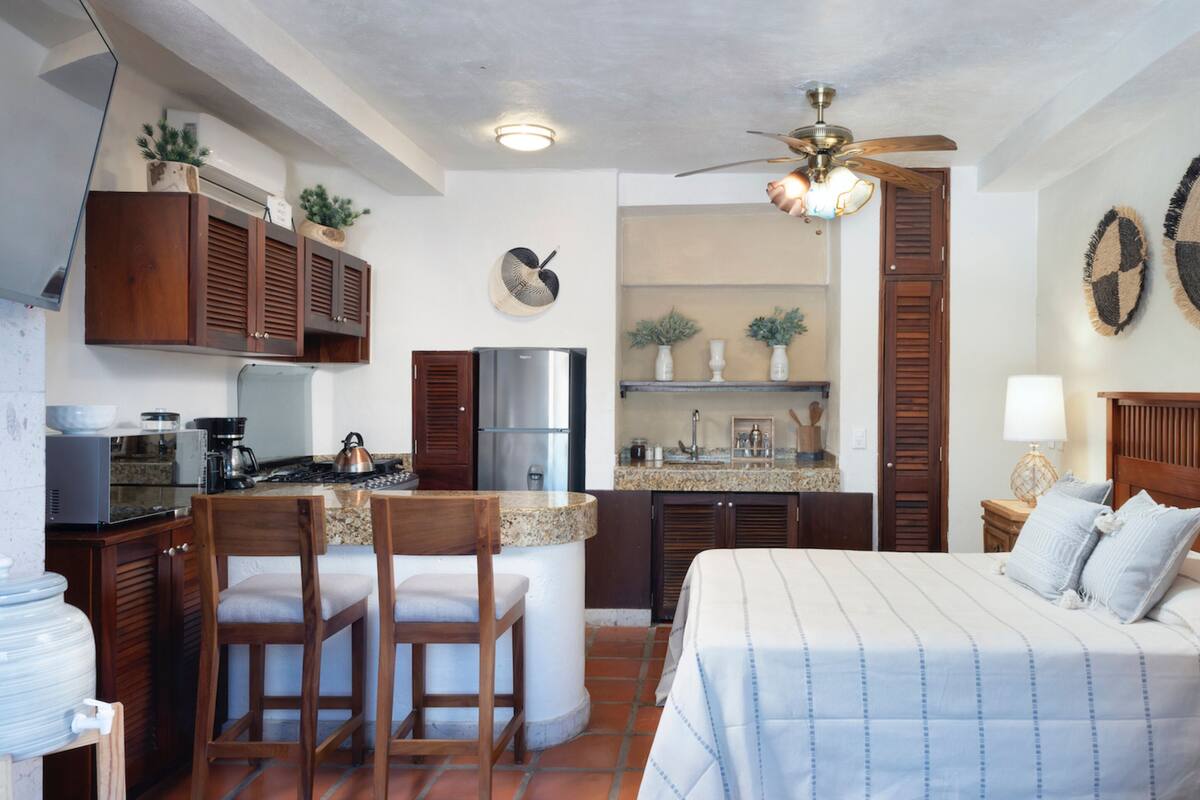 Cozinha compacta com geladeira, micro-ondas e cafeteira. Há uma área de refeições com cadeiras e uma pequena ilha. O quarto possui uma cama com cabeceira de madeira e uma mesa de cabeceira com abajur.