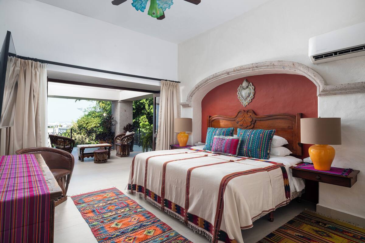 Quarto com cama, mesa de cabeceira, cadeira, mesa e vista para o verde lá fora.