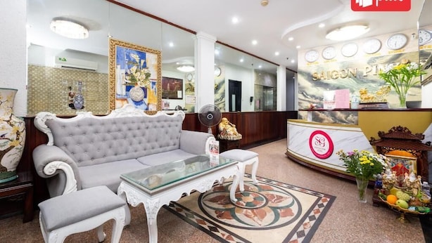 Um saguão de hotel com um sofá cinza, uma mesa de centro de vidro e um balcão de recepção com um relógio e uma placa com a inscrição "SAIGON PIN".