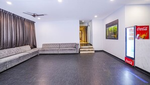 Um lobby moderno de hotel com um sofá modular cinza, um ventilador de teto, um refrigerador de Coca-Cola e uma escada que leva a outra área.