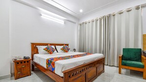 Cama bem arrumada com estrutura de madeira e almofadas coloridas, mesa de cabeceira com um telefone e uma cadeira verde num quarto bem iluminado com paredes brancas e cortina.