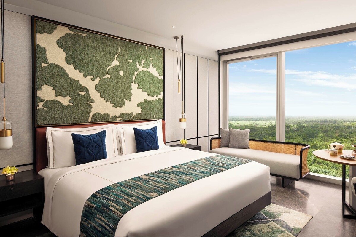 Quarto de hotel moderno com uma cama grande, área de estar com vista e uma mesa pequena com chá e acompanhamentos.