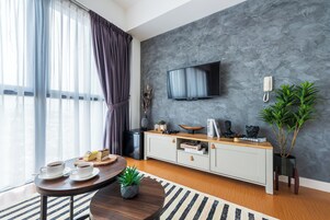 Uma sala de estar moderna com uma TV de tela plana fixada em uma parede cinza texturizada, uma mesa de centro de madeira com xícaras e uma sobremesa, um armário branco com objetos decorativos e um vaso de planta.