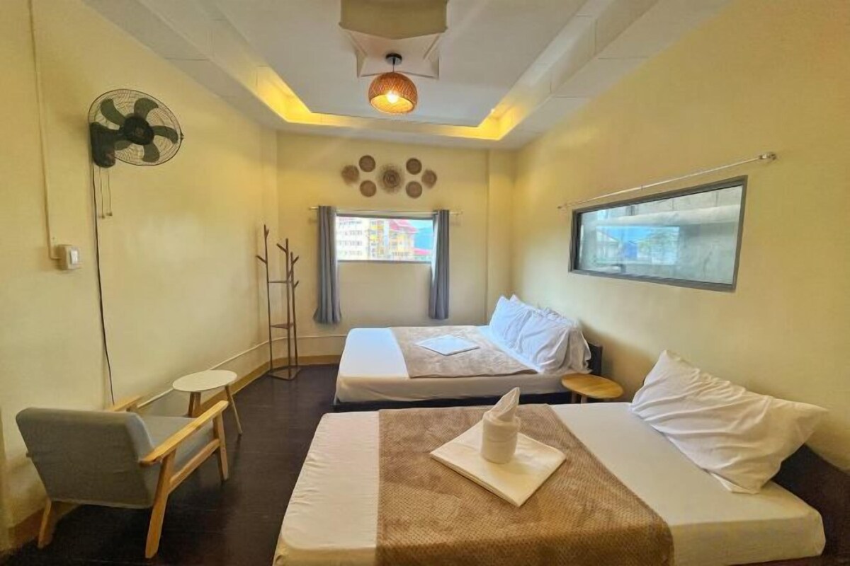 Quarto de hotel com duas camas, uma escrivaninha, uma cadeira, um ventilador e janela com vista.
