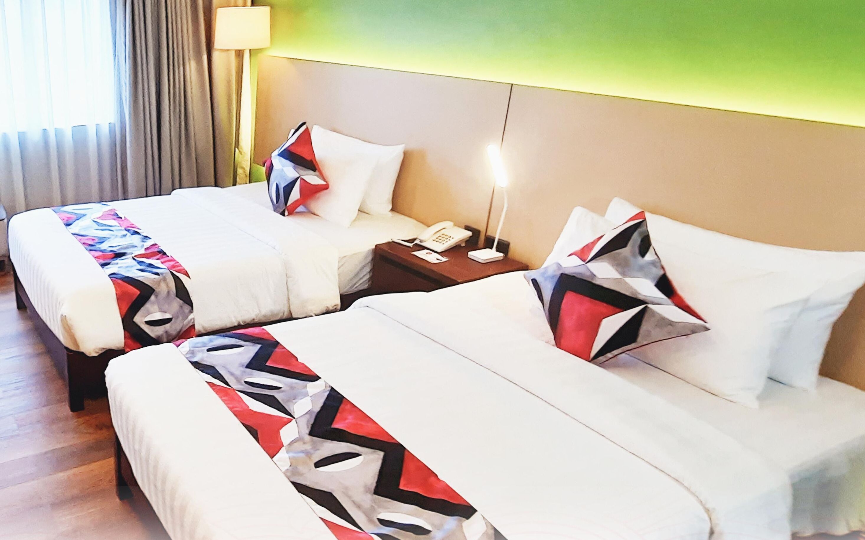 Duas camas de hotel com lençóis brancos e cobertores de padrão geométrico, uma mesa de cabeceira com luminária e um telefone.