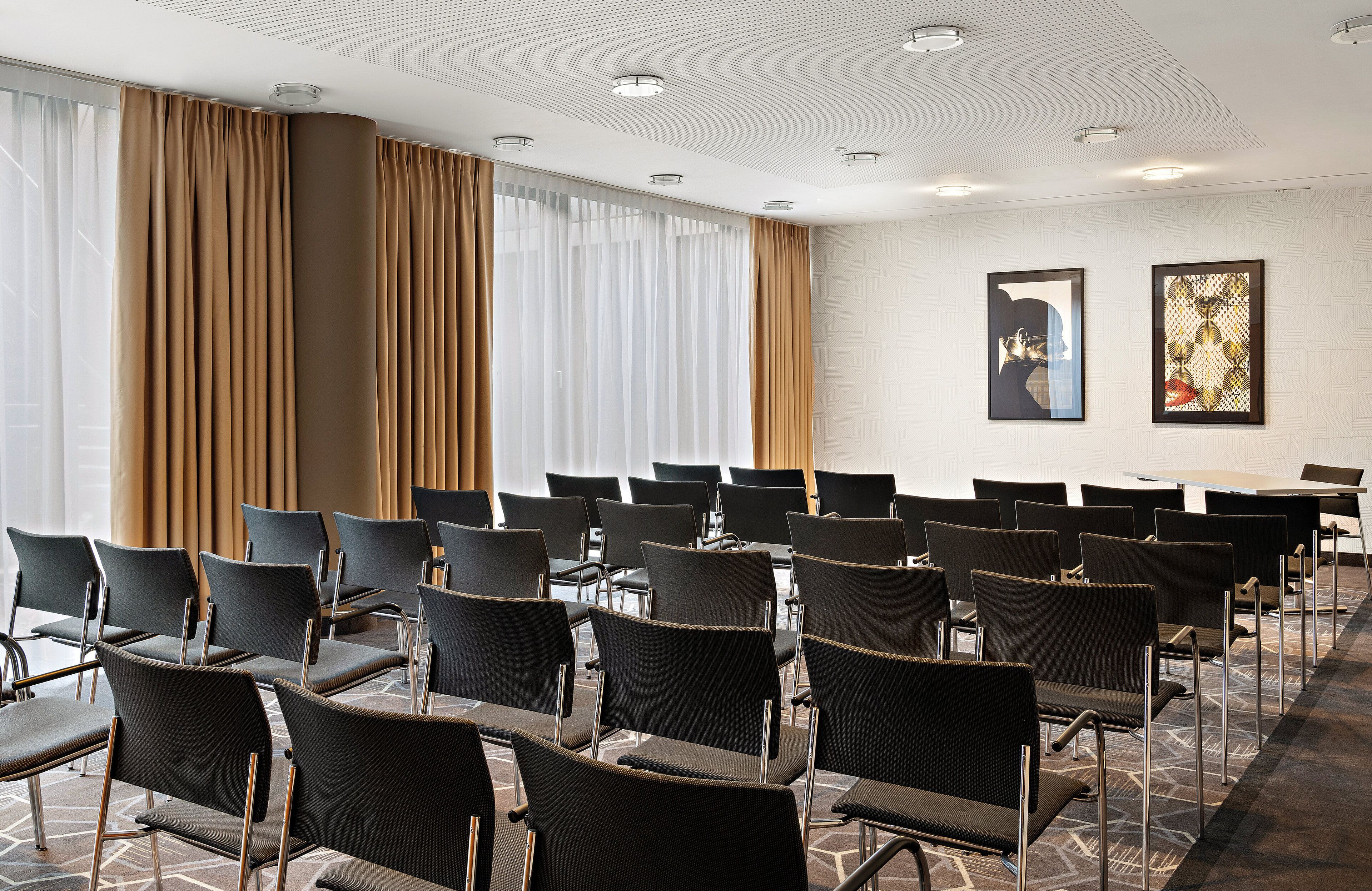Sala de conferências com fileiras de cadeiras, uma mesa e grandes janelas com cortinas.