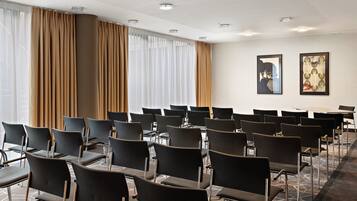 Sala de conferências com fileiras de cadeiras, uma mesa e grandes janelas com cortinas.