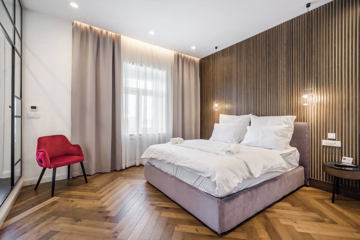 Quarto moderno com uma cama grande, uma cadeira vermelha, piso de madeira e luminária fixada na parede.