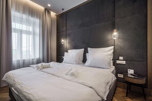 Quarto de hotel moderno com uma cama grande, roupa de cama branca, cabeceira escura e mesa de cabeceira com um controle remoto.