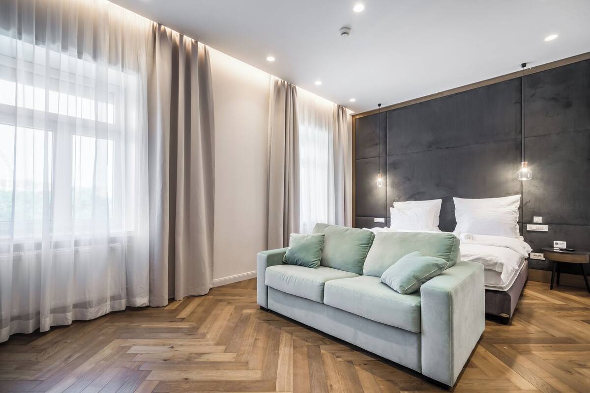 Quarto moderno com um sofá de cor clara, uma cama com roupa de cama branca e uma janela grande com cortinas translúcidas.